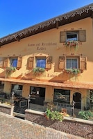 Um edifício com uma placa que diz "Room and Breakfast Ladin". Apresenta fachada de tijolos, persianas de madeira e cestos de flores pendurados.