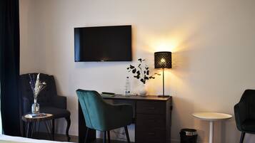 Quarto de hotel com cama, escrivaninha, duas cadeiras, televisão, luminária e mesinha de centro com um vaso e livros.