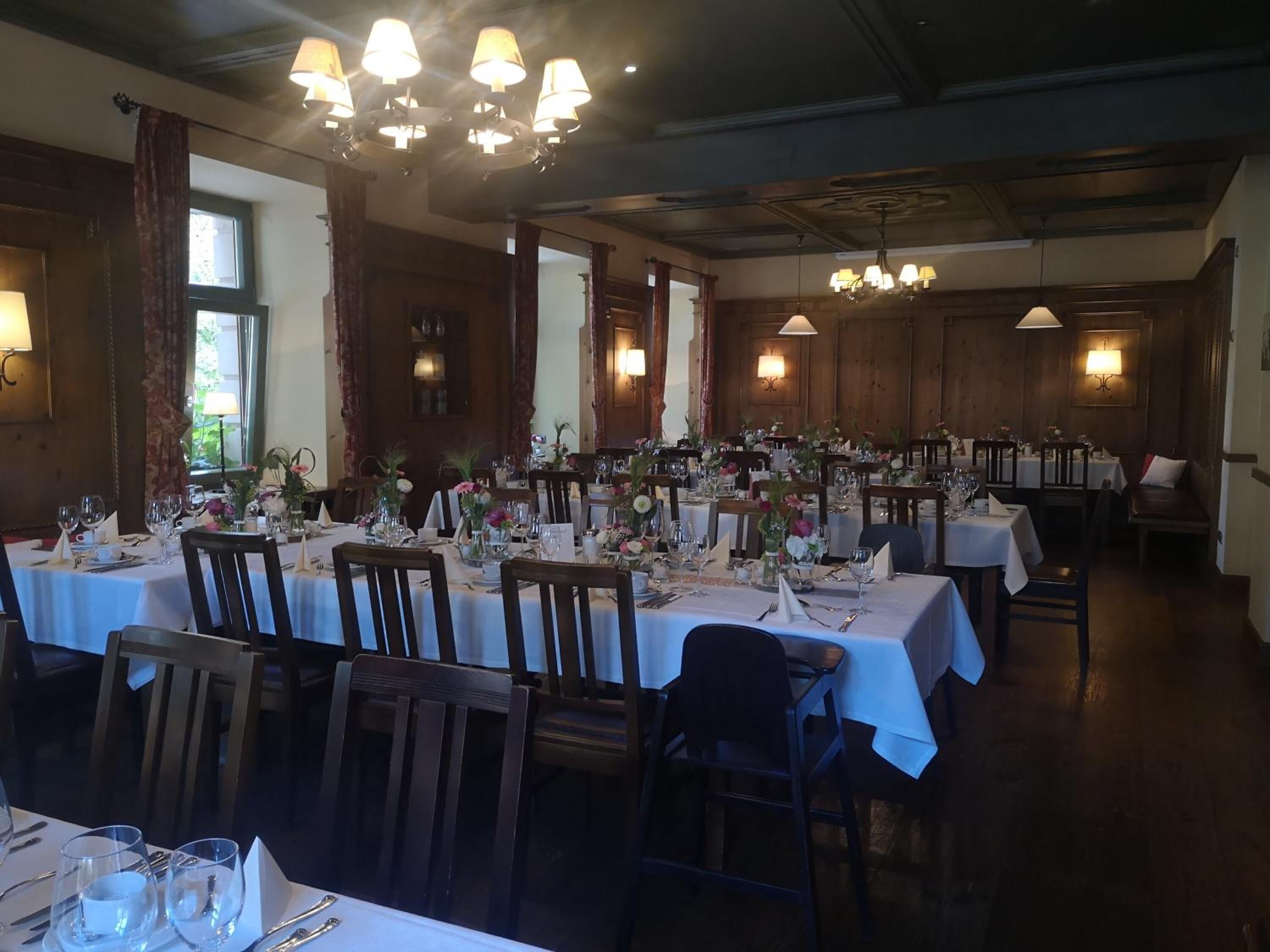 Uma sala de jantar com várias mesas preparadas para o serviço, cadeiras de madeira e lustres.