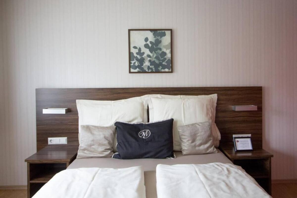 Cama bem arrumada com lençóis brancos e almofada escura com monograma, acompanhada por duas mesas de cabeceira com luminárias.