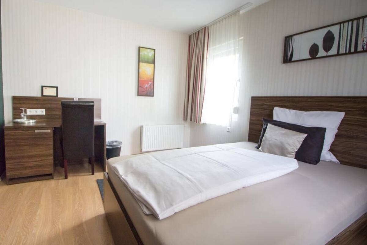 Quarto de hotel com cama, escrivaninha e cadeira. Há uma janela com cortinas, um radiador e um quadro na parede.