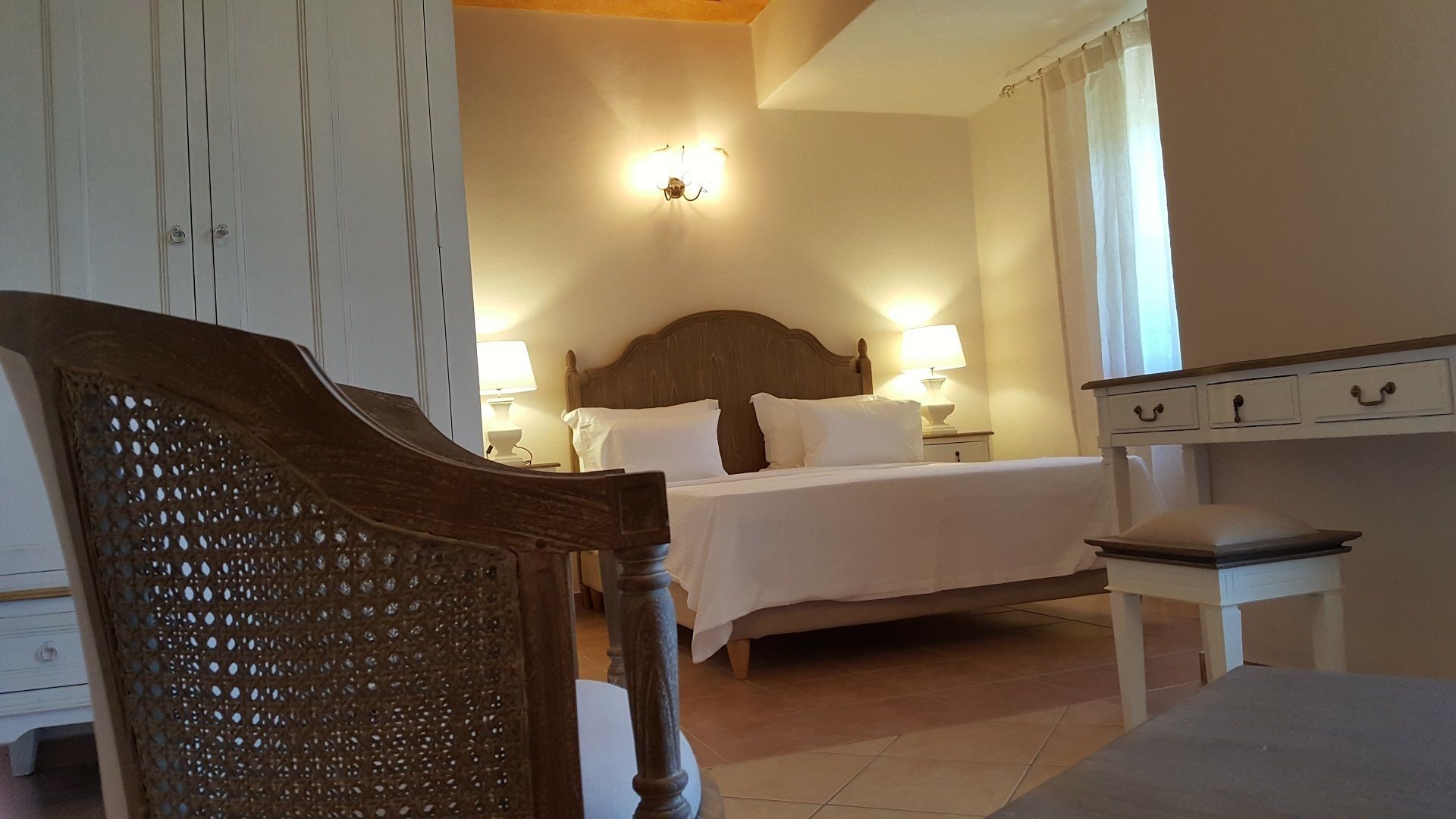 Quarto de hotel com cama de madeira, duas luminárias de cabeceira, mesa de apoio, uma cadeira e uma pequena mesa.