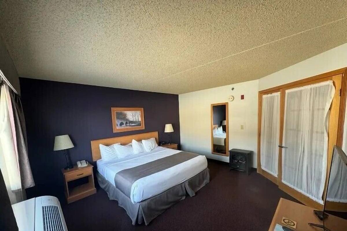 Quarto de hotel com uma cama grande, uma mesinha de cabeceira, uma escrivaninha com cadeira, um espelho e uma janela com cortinas.