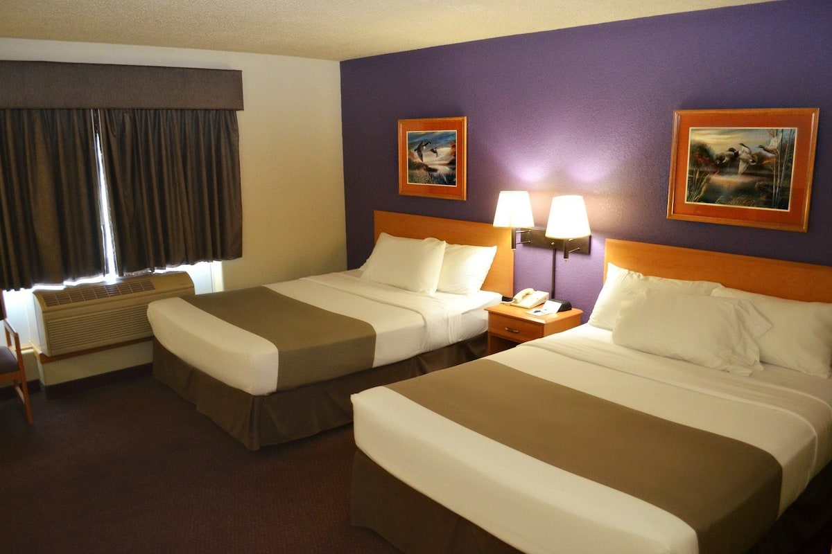 Quarto de hotel com duas camas, uma janela com cortinas, ar condicionado, mesa de cabeceira com abajur e duas molduras na parede.