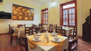 Uma sala de jantar com várias mesas preparadas para hóspedes, janelas amplas e um lustre.