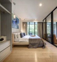 Quarto moderno com uma cama grande, mesa de cabeceira, luminária pendente e vista para o verde através de janelas amplas.
