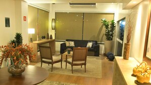 Uma sala de estar moderna com sofá, cadeiras, uma mesa de centro e um espaço de jantar com mesa e cadeiras. Há uma janela grande com persianas, uma planta e um vaso decorativo.