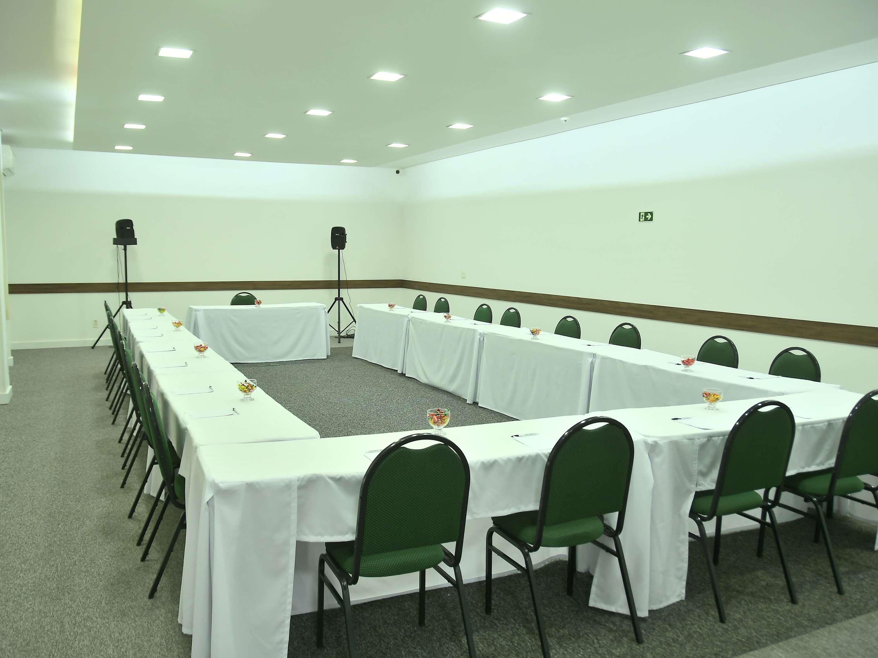 Uma sala de conferência com uma mesa comprida, cadeiras verdes, toalhas de mesa brancas e iluminação no teto.