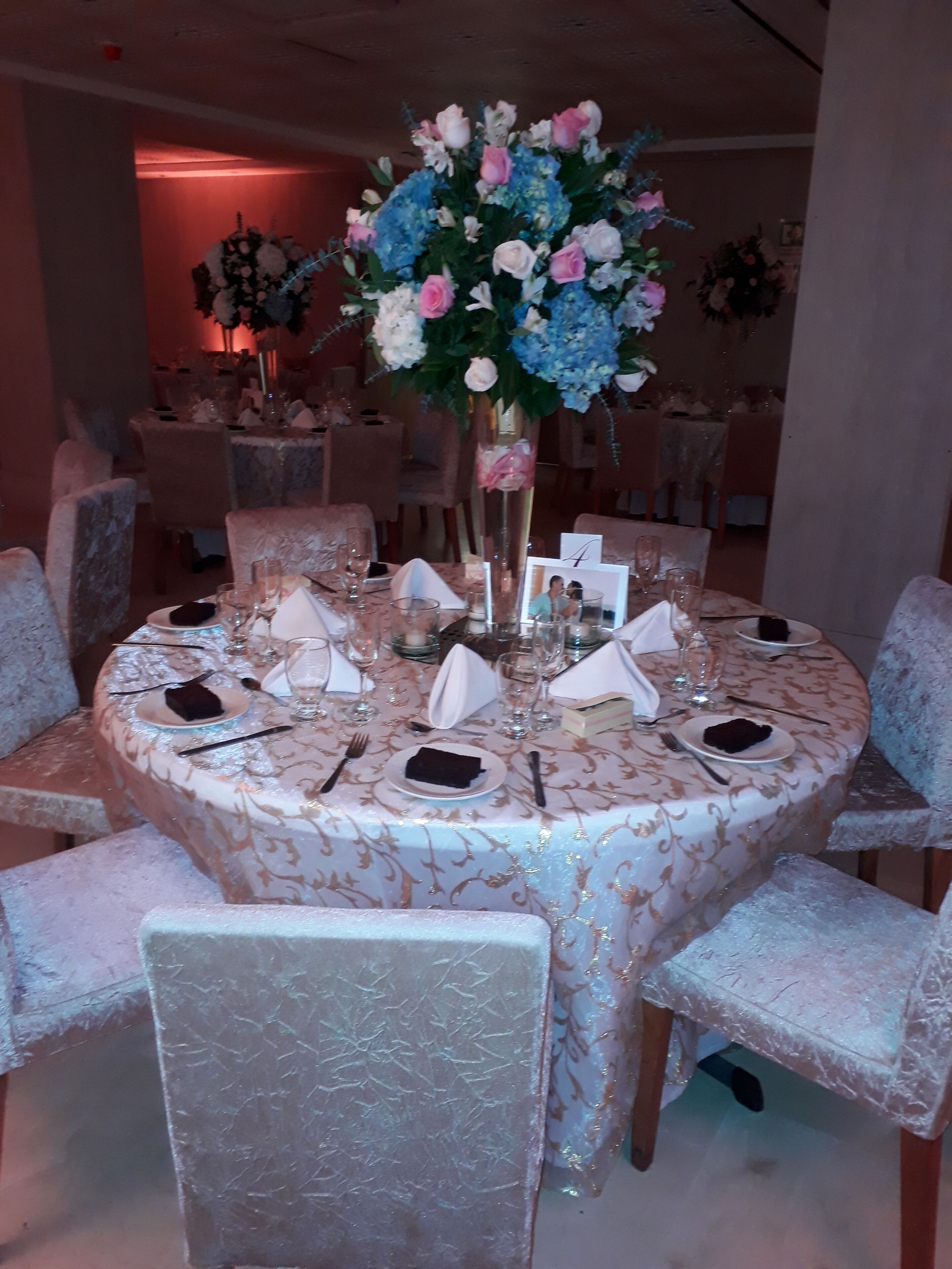 Uma mesa de jantar redonda com toalha branca, rodeada por cadeiras com tecido texturizado. Um vaso alto com flores está posicionado no centro.