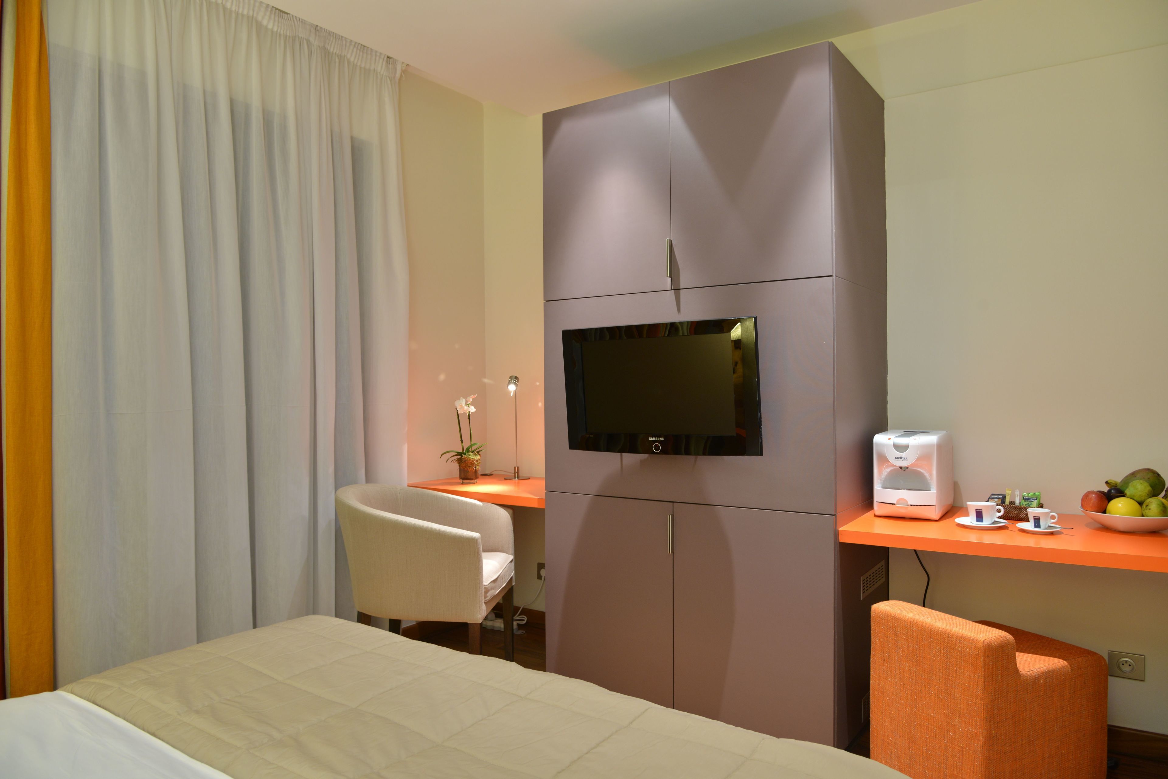 Quarto de hotel com cama, escrivaninha com cadeira, televisão e pequena mesa com fruteira e cafeteira.