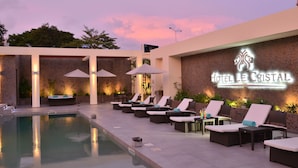 Uma área de piscina de hotel com espreguiçadeiras e guarda-sóis, um muro de tijolos com uma placa indicando "Hotel Le Cristal", e um céu limpo.