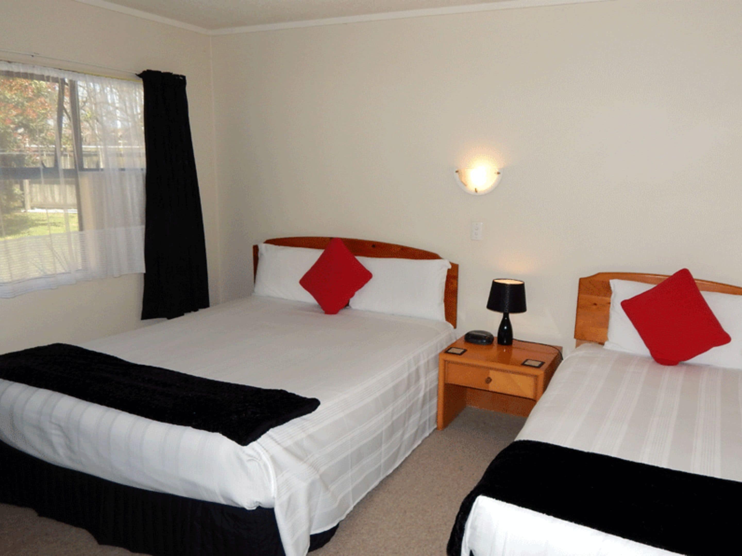 Quarto de hotel com duas camas, uma mesa de cabeceira com abajur, uma janela com cortinas e um candeeiro fixo na parede.