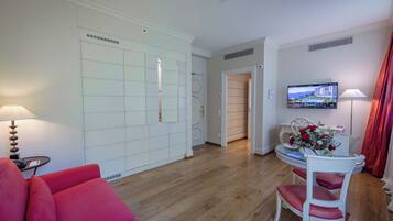 Quarto de hotel com piso de madeira, sofá vermelho, mesa de jantar branca com cadeiras, TV na parede e uma janela grande.