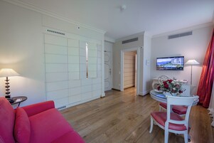 Quarto de hotel com piso de madeira, sofá vermelho, mesa de jantar branca com cadeiras, TV na parede e uma janela grande.