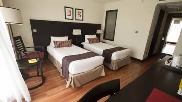 Quarto de hotel com duas camas de solteiro, uma escrivaninha com cadeira, uma luminária e uma janela.