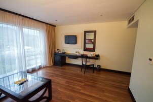 Quarto de hotel com chão de madeira, uma mesa de vidro com um controle remoto, uma escrivaninha com uma cadeira, uma televisão na parede e uma janela com cortinas.