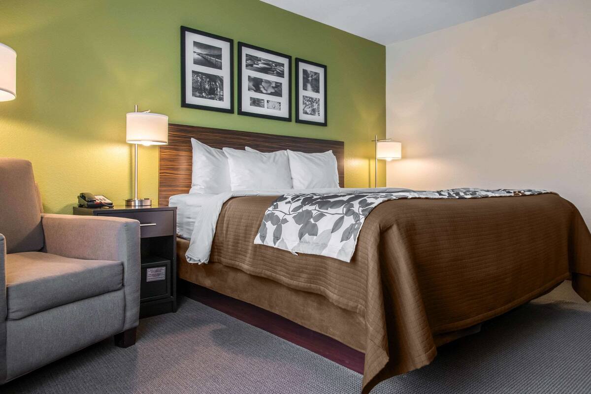 Quarto de hotel com cama, uma cadeira, mesa de cabeceira e luminárias fixadas na parede.