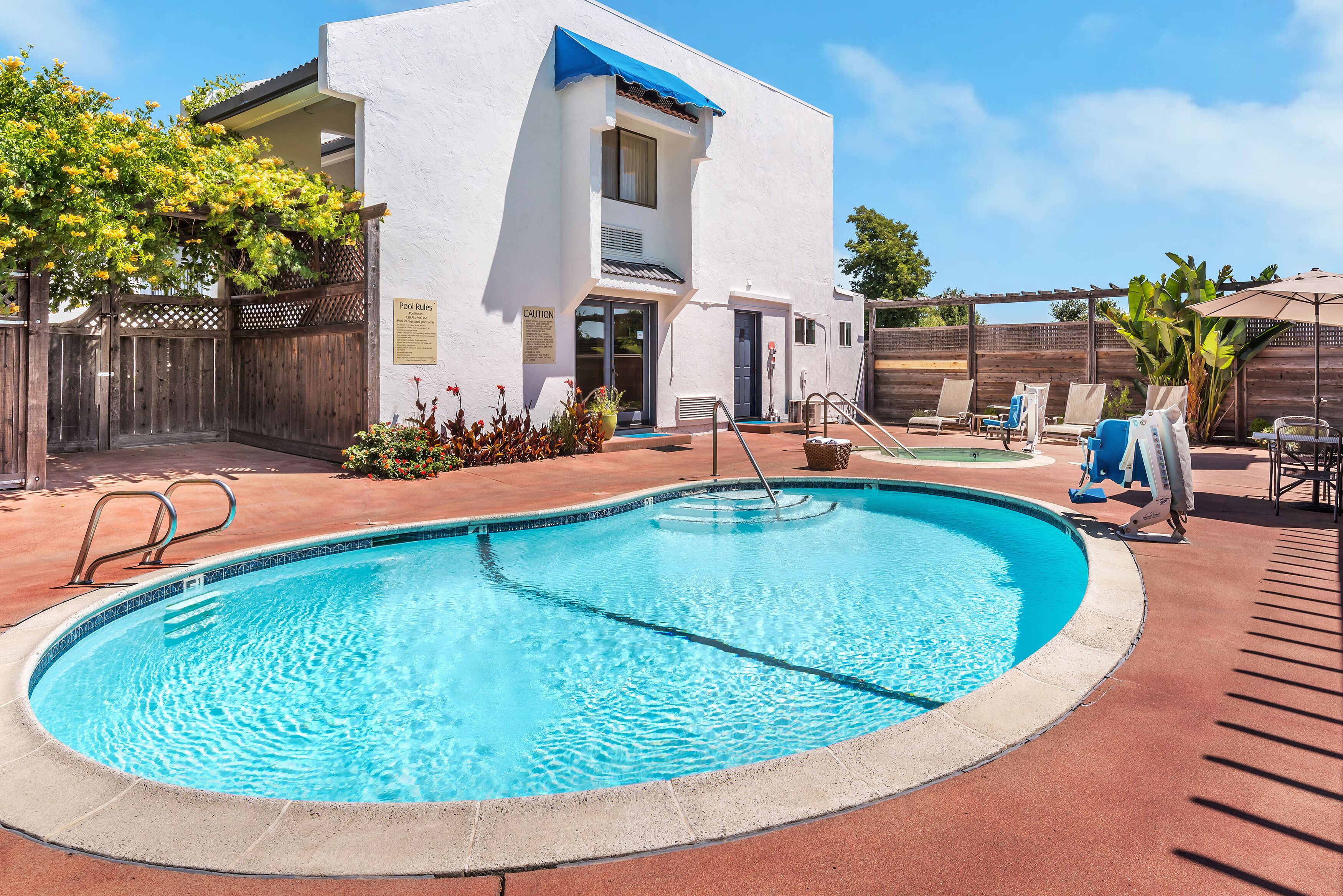 Piscina com deck de madeira, um edifício branco com toldo azul e uma área cercada com cadeiras de descanso.