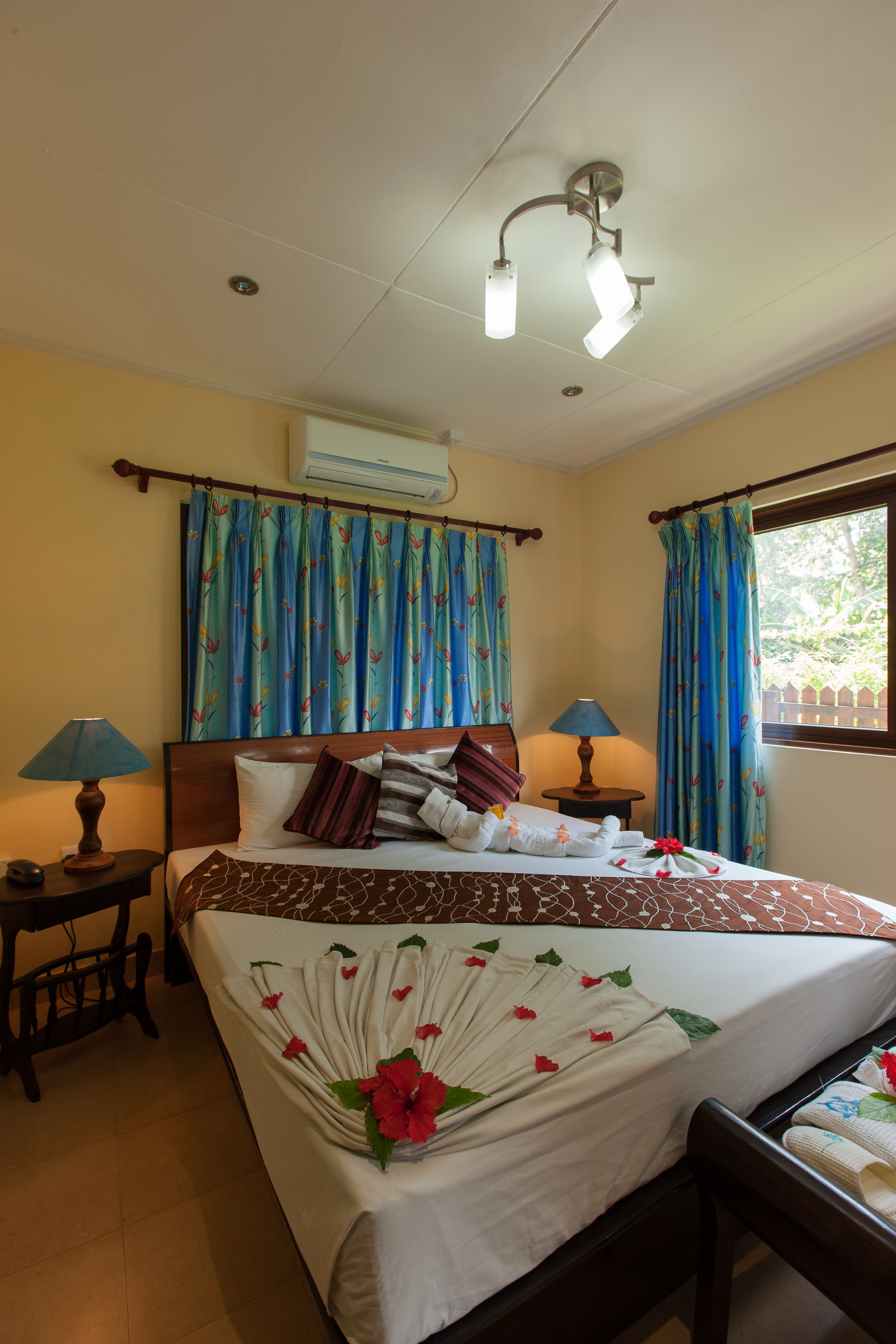 Quarto de hotel com cama, duas mesas de cabeceira com abajures, uma janela com cortinas e um aparelho de ar condicionado.