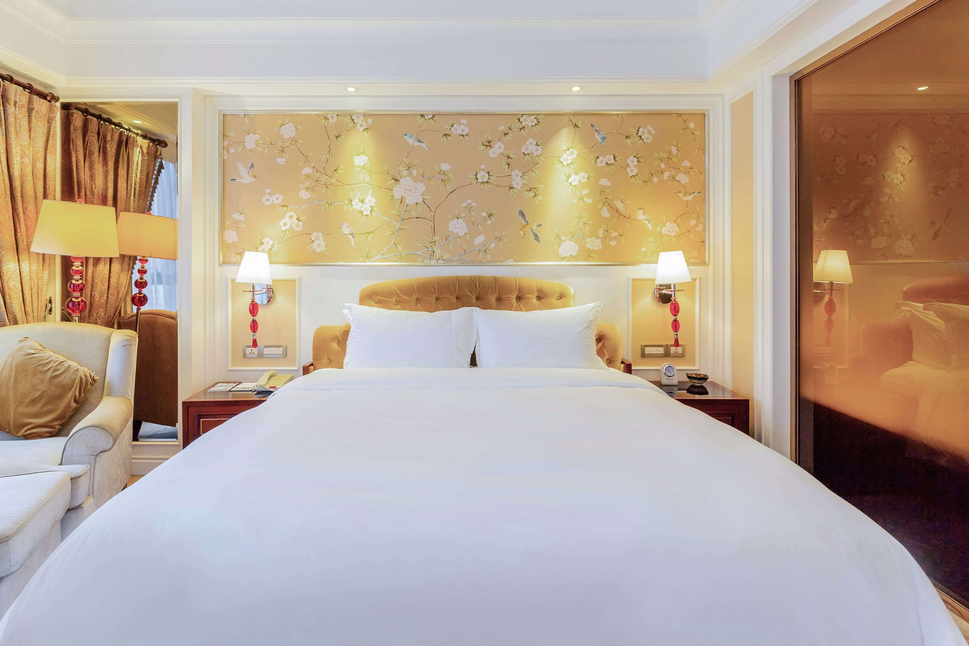 Quarto de hotel com uma cama grande, duas mesas de cabeceira com luminárias, uma cadeira e uma parede decorativa com padrões florais.