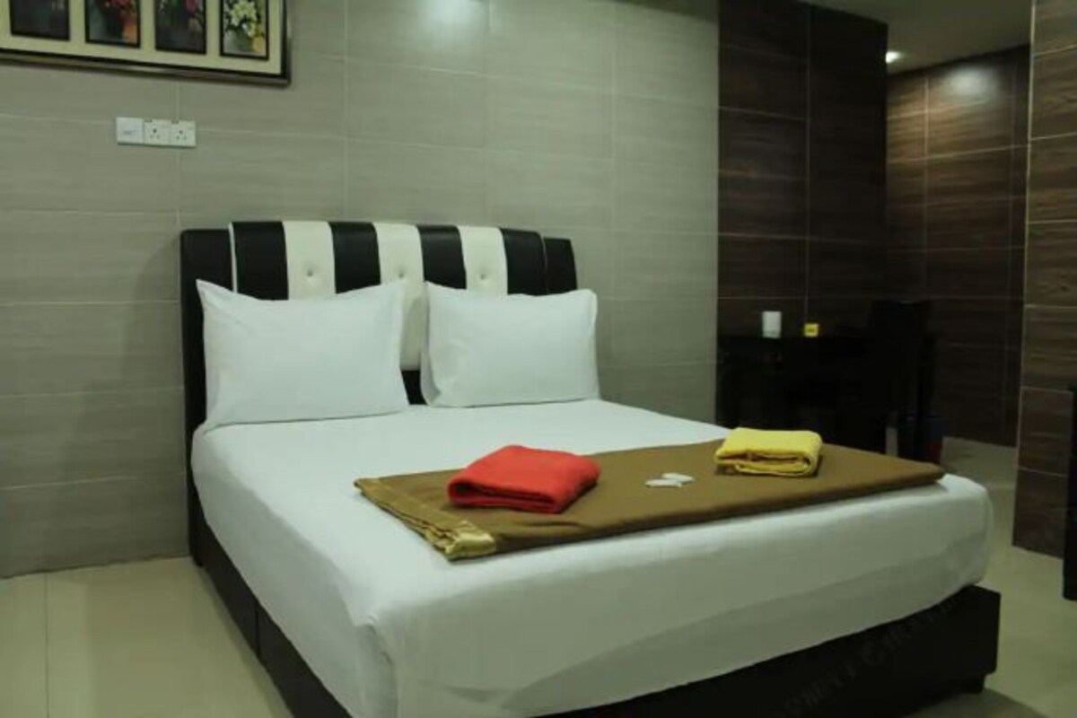 Quarto de hotel com uma cama, almofadas brancas, manta marrom e vermelha, e uma mesinha com uma garrafa.