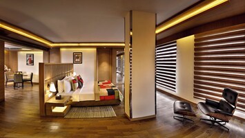 Quarto de hotel moderno com piso de madeira, cama com almofadas coloridas, escrivaninha com luminária e uma cadeira.