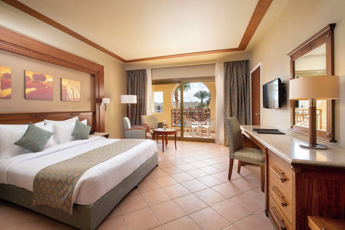 Quarto de hotel com uma cama grande, uma escrivaninha, uma cadeira, uma televisão e uma varanda com vista.