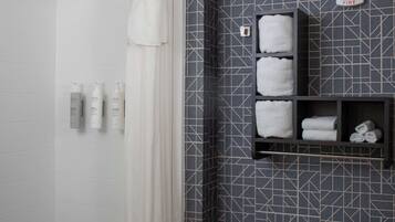 Casa de banho com parede revestida a mosaico branco, cortina de chuveiro e prateleira com toalhas e produtos de higiene pessoal.