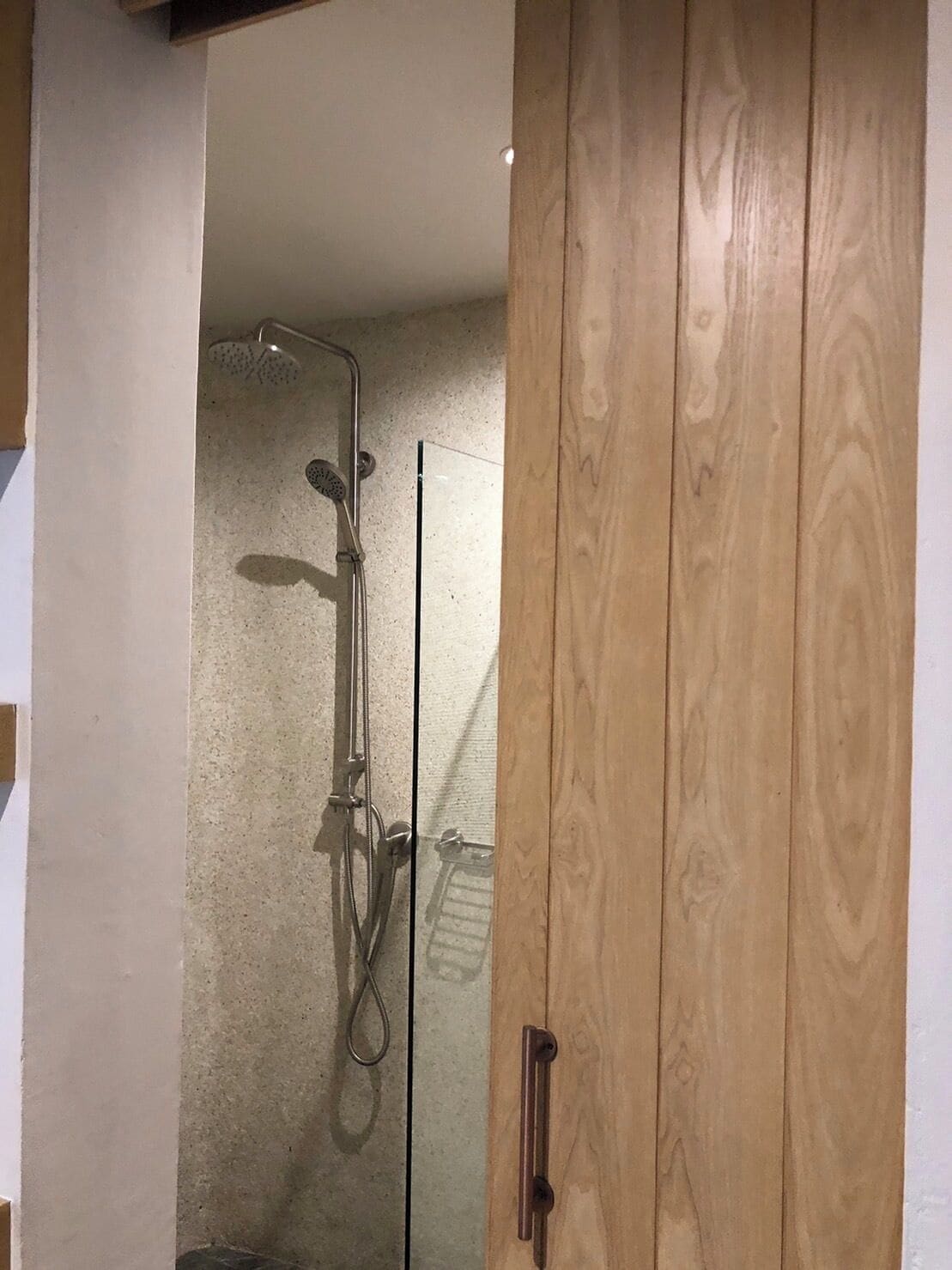 Uma porta de madeira com puxador de metal, dando acesso a um espaço de chuveiro com porta de vidro e chuveiro com cabeça metálica.