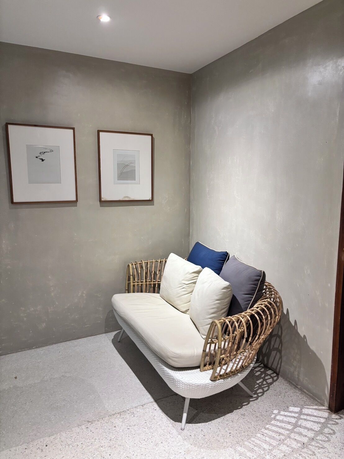 Quarto de canto com um sofá de vime, duas fotos emolduradas na parede e piso de concreto.