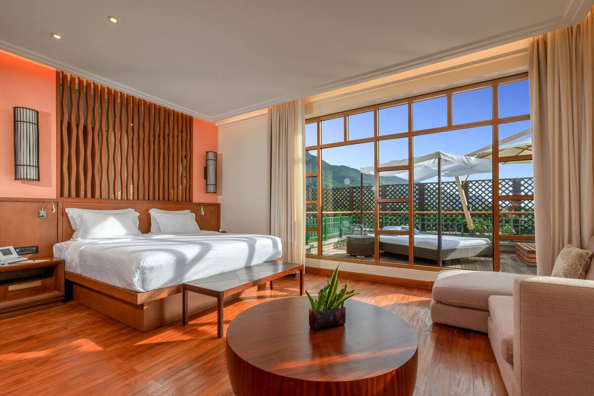 Quarto de hotel moderno com uma cama grande, um espaço de estar, uma mesa de centro redonda e vista para uma varanda com mobiliário de exterior.