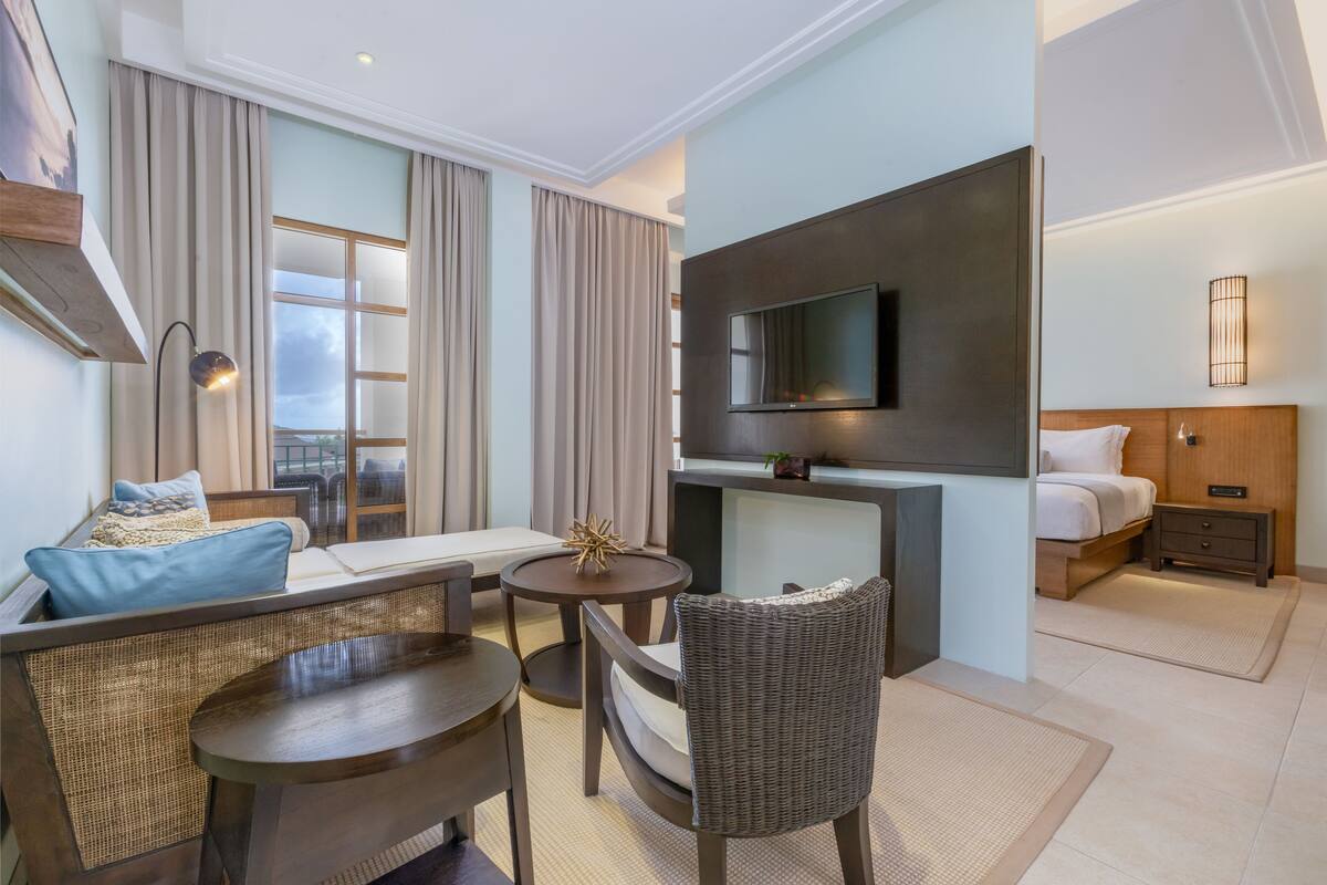 Quarto de hotel moderno com sofá, mesa de jantar e cadeiras, televisão na parede, cama e pequena mesa de apoio.