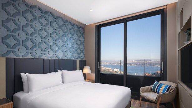 Quarto de hotel moderno com uma cama grande, vista para a cidade e um painel decorativo na parede.