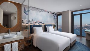 Quarto de hotel moderno com uma cama grande, banheiro com pia e espelho, e vista para a cidade através de grandes janelas.