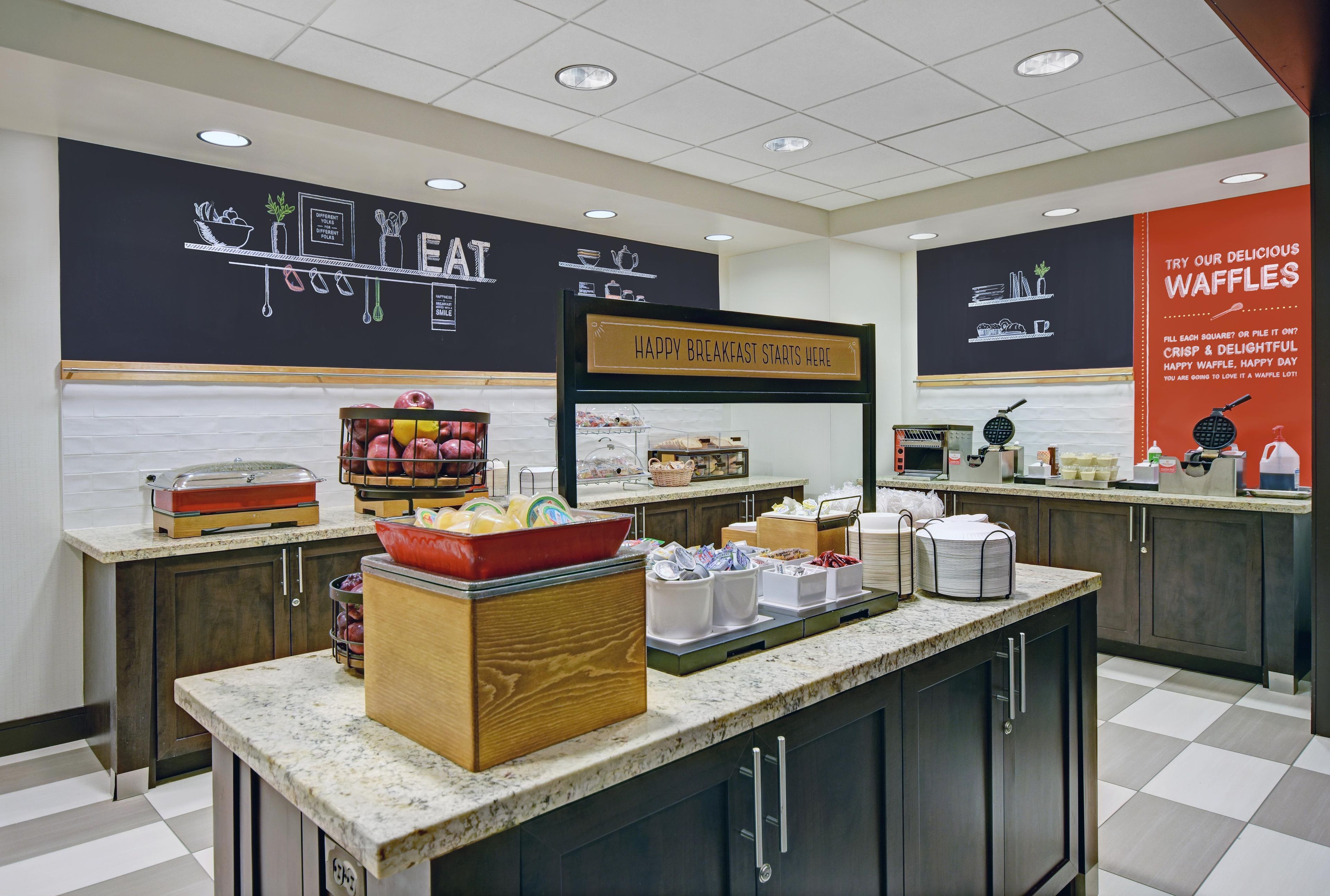 Espaço de buffet de café da manhã com placa indicando o início do serviço, diversas opções de alimentos e uma estação de waffles.