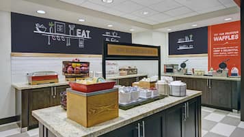 Espaço de buffet de café da manhã com placa indicando o início do serviço, diversas opções de alimentos e uma estação de waffles.