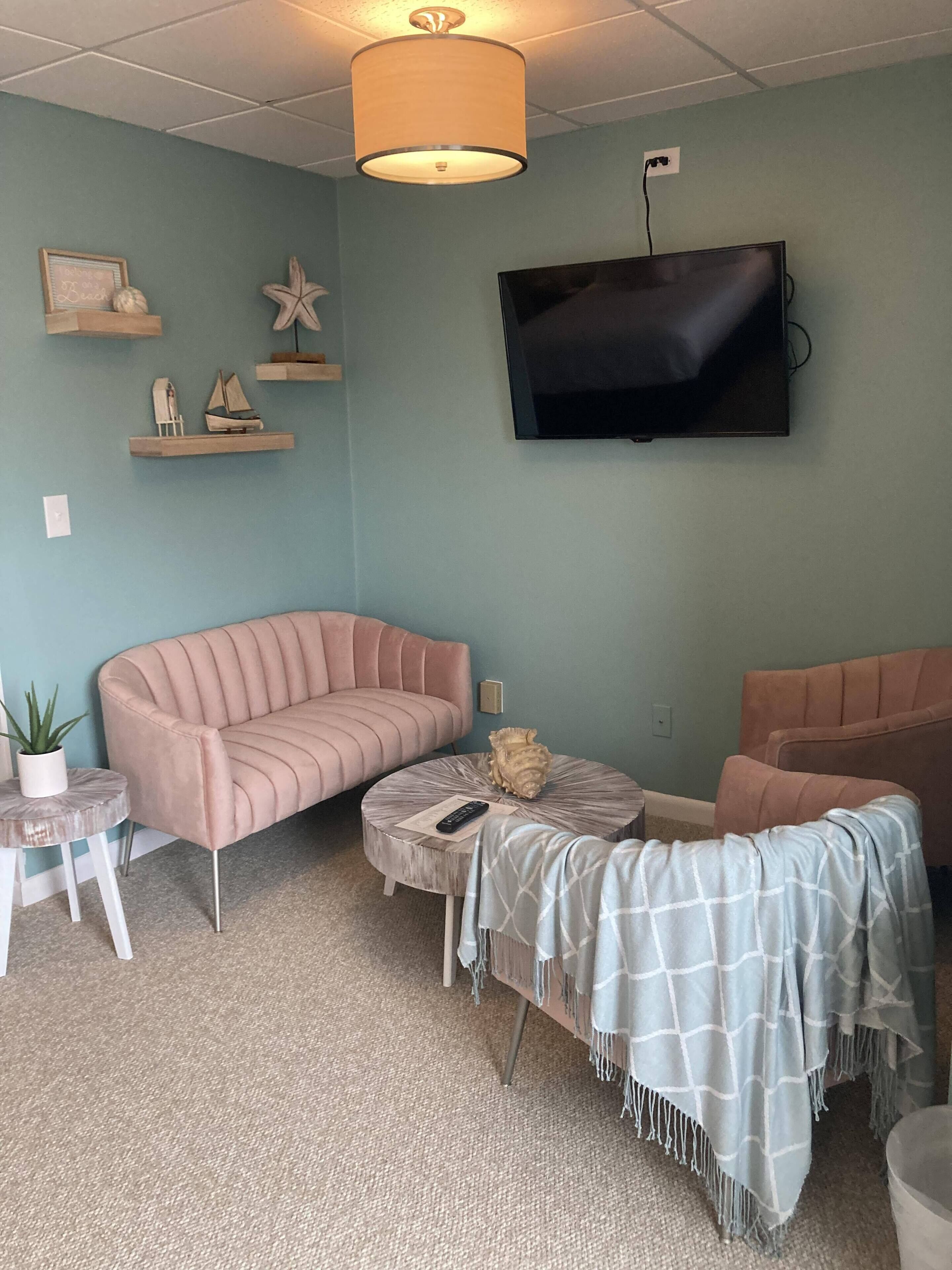 Uma sala de estar com um sofá rosa, uma mesa de centro redonda, uma TV fixada na parede e uma pequena mesa lateral com uma planta.