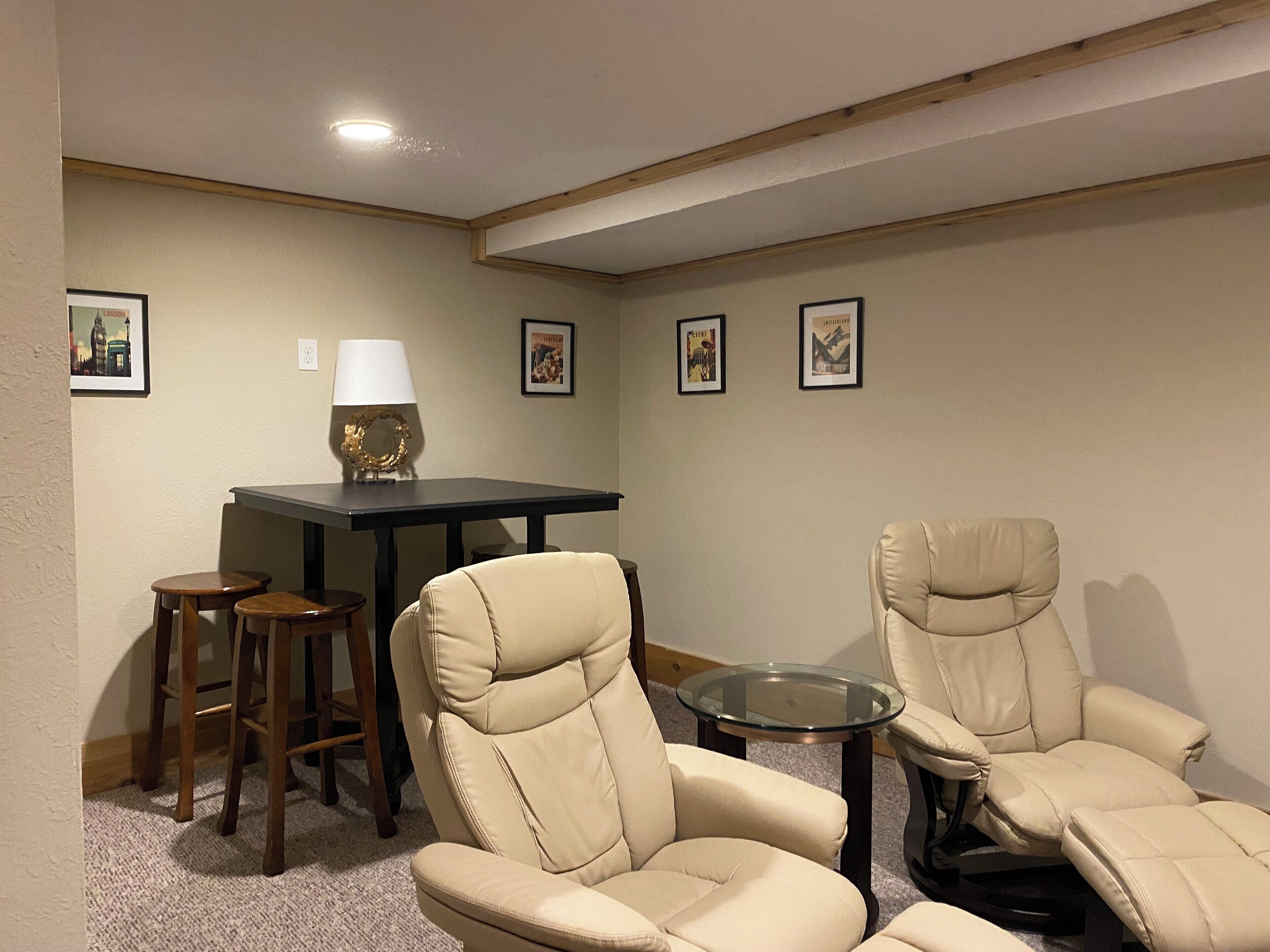 Quarto com duas poltronas de couro bege, uma mesa redonda de vidro e uma mesa de madeira com abajur.