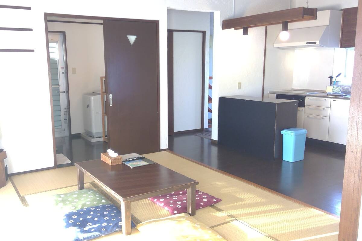 Uma sala de estar com televisão, uma mesa de madeira, esteiras de tatame e uma cozinha com pia e armários.