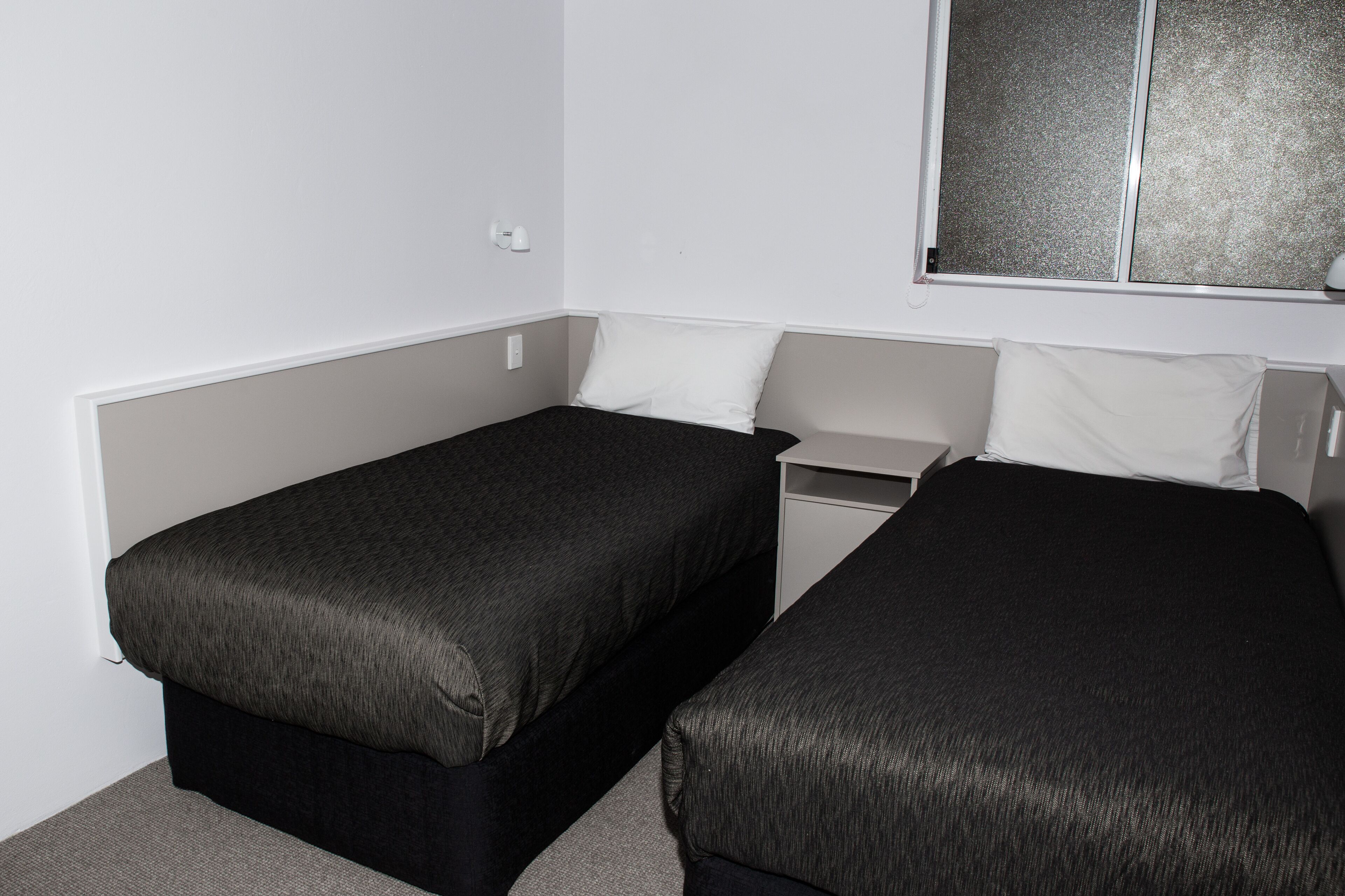 Quarto pequeno e individual com duas camas de solteiro, mesa de cabeceira e janela com persianas.
