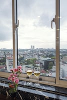 Uma janela com vista para a paisagem urbana, um vaso com flores cor-de-rosa e duas taças de vinho branco no parapeito.