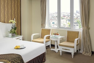 Quarto de hotel com uma cama, duas cadeiras listradas, uma mesa pequena com uma cesta de frutas e vista para a cidade pela janela.