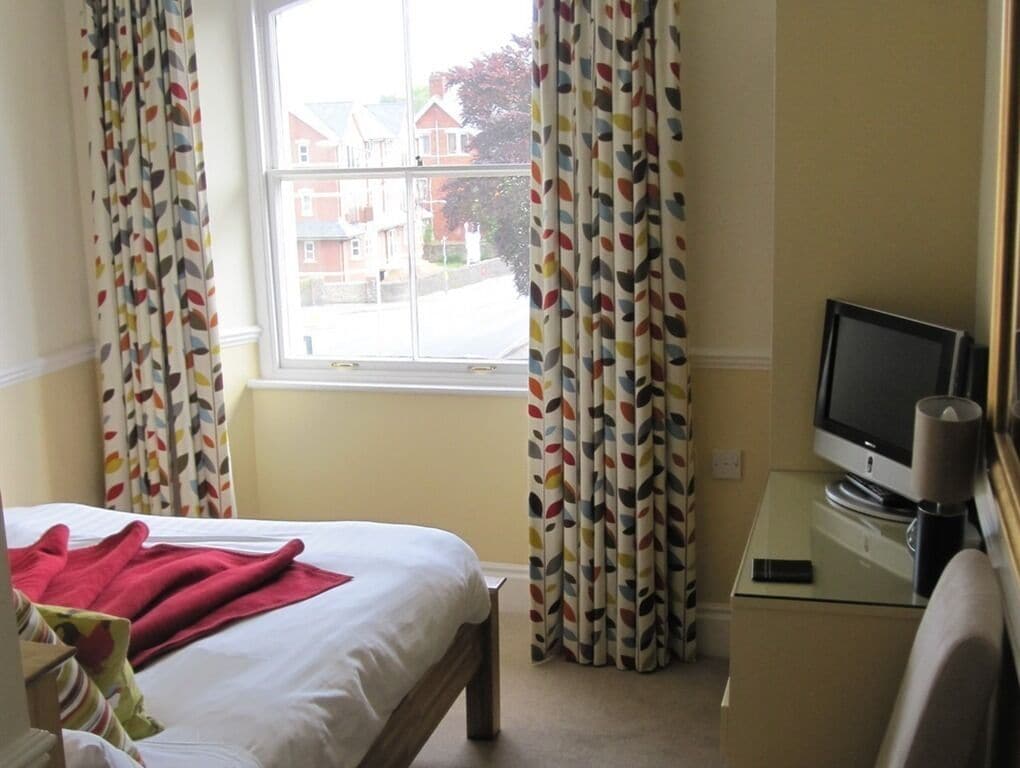 Quarto de hotel com uma cama, uma escrivaninha com computador e uma janela com cortinas.