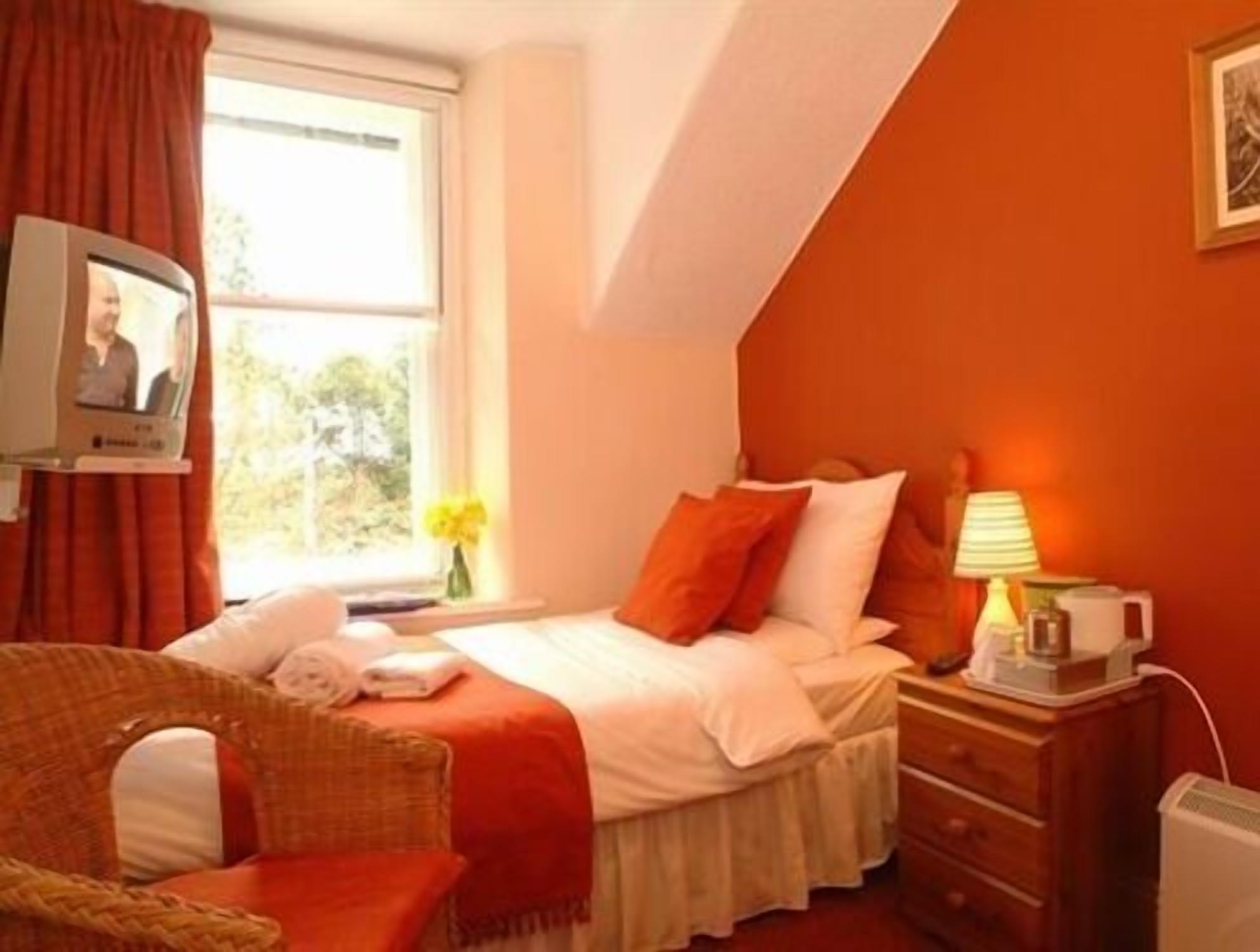 Quarto de hotel com cama, televisão na parede, janela com cortinas, mesa de cabeceira com abajur e uma cadeira.