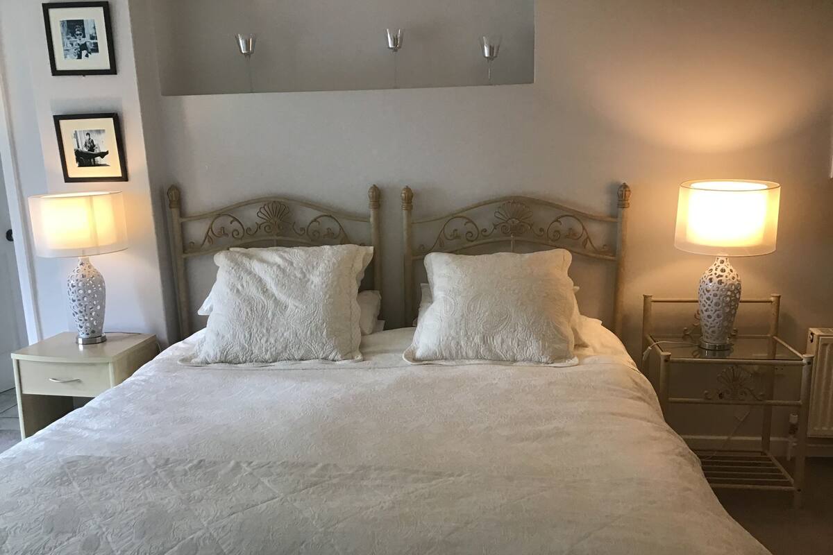 Cama bem arrumada com duas almofadas, mesa de cabeceira com abajur e luminária na parede.