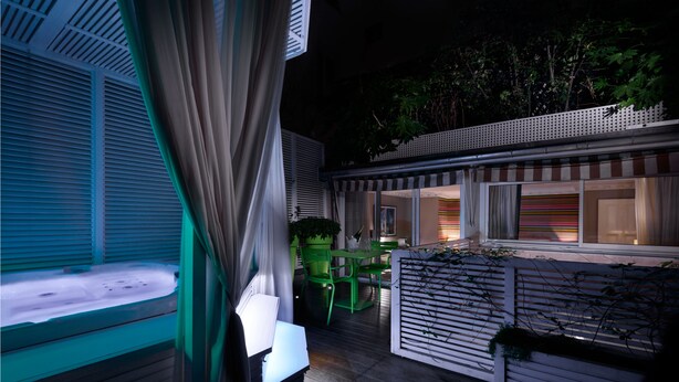 Uma área externa coberta com deck de madeira, cadeiras verdes, uma mesa e um banco.