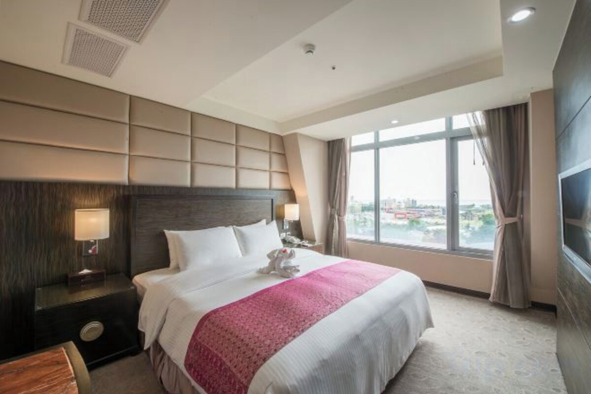 Quarto de hotel com uma cama grande, mesa de cabeceira, televisão e vista para a cidade.