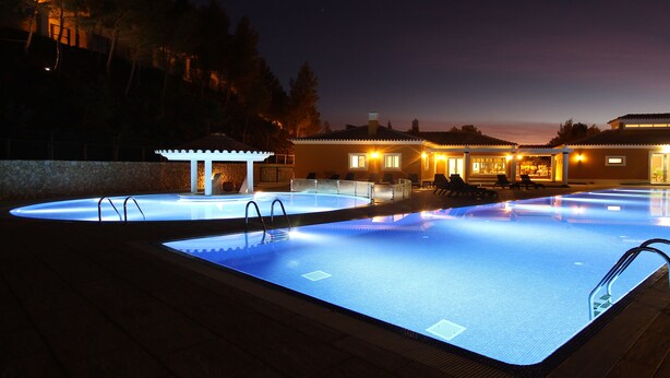 Uma área de piscina com duas piscinas, espreguiçadeiras e um espaço de estar coberto ao entardecer.