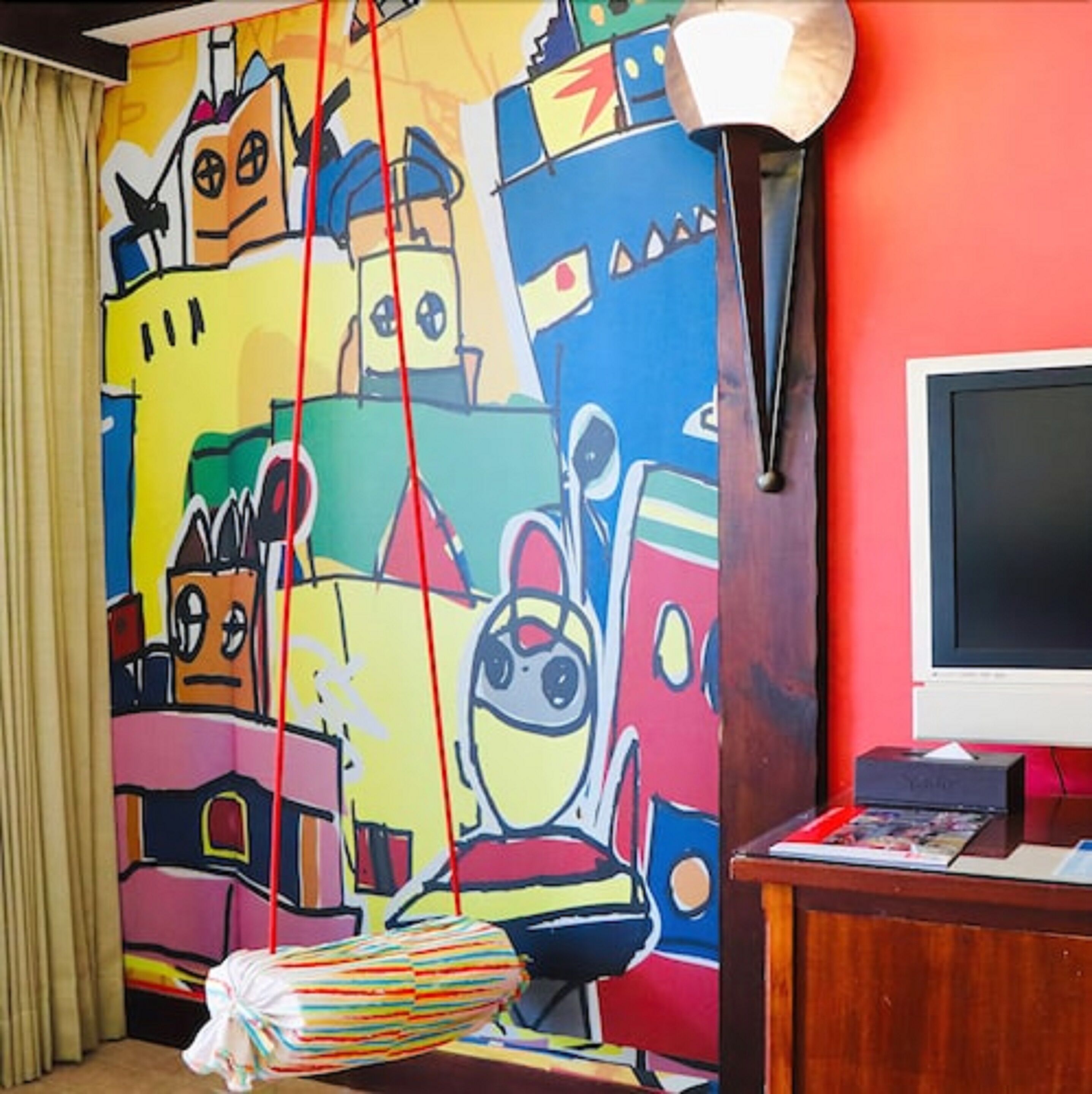 Quarto com um mural abstrato colorido na parede, uma escrivaninha com um computador e um almofada listrada no chão.