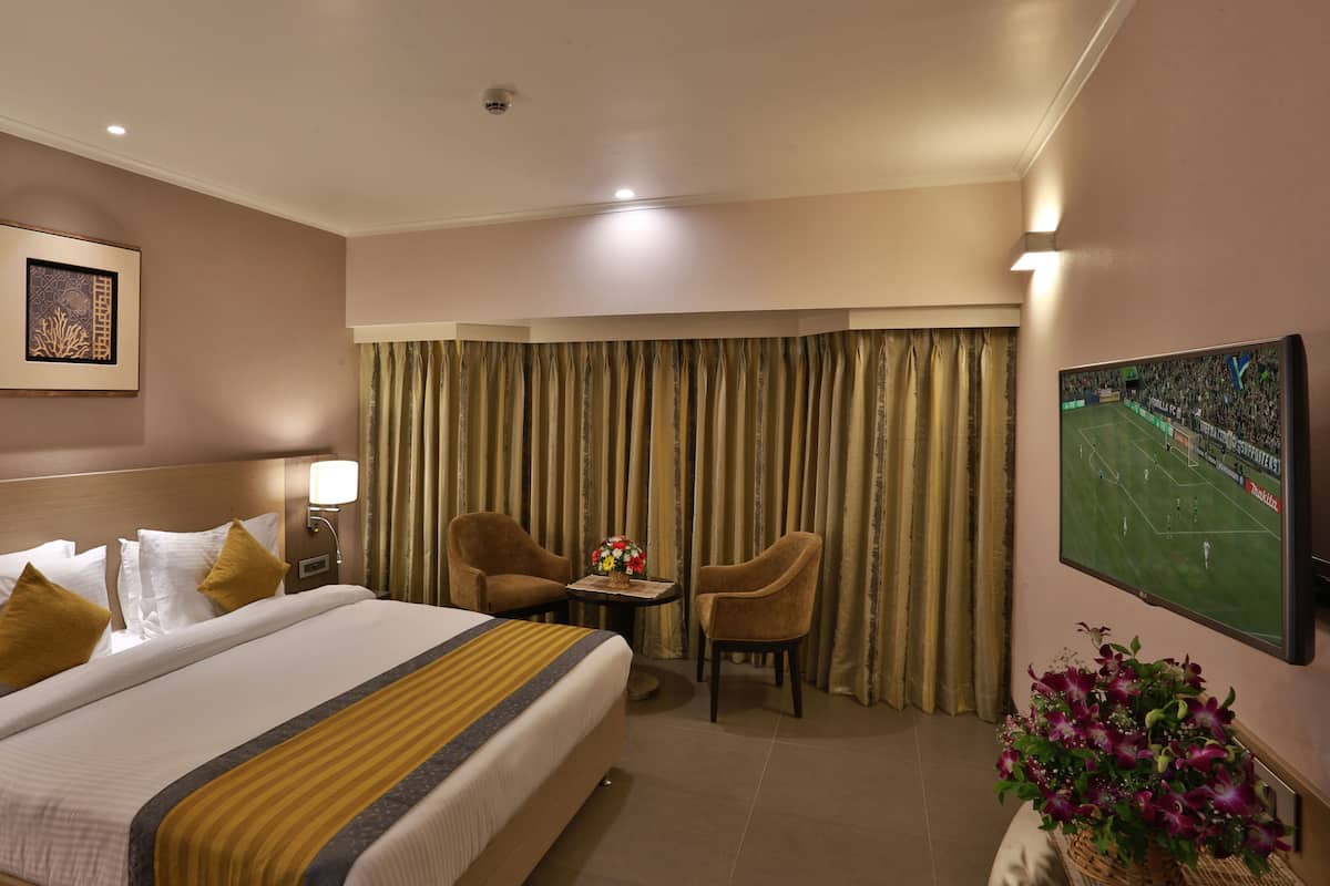 Quarto de hotel com uma cama, duas cadeiras, uma mesinha com um vaso, um quadro na parede e uma TV exibindo um jogo de futebol.